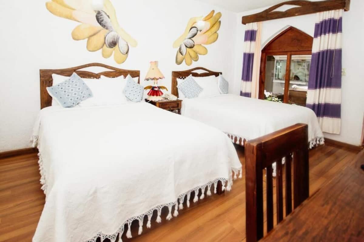 Quarto com duas camas de madeira, uma mesa de cabeceira, uma luminária e uma janela com cortinas.