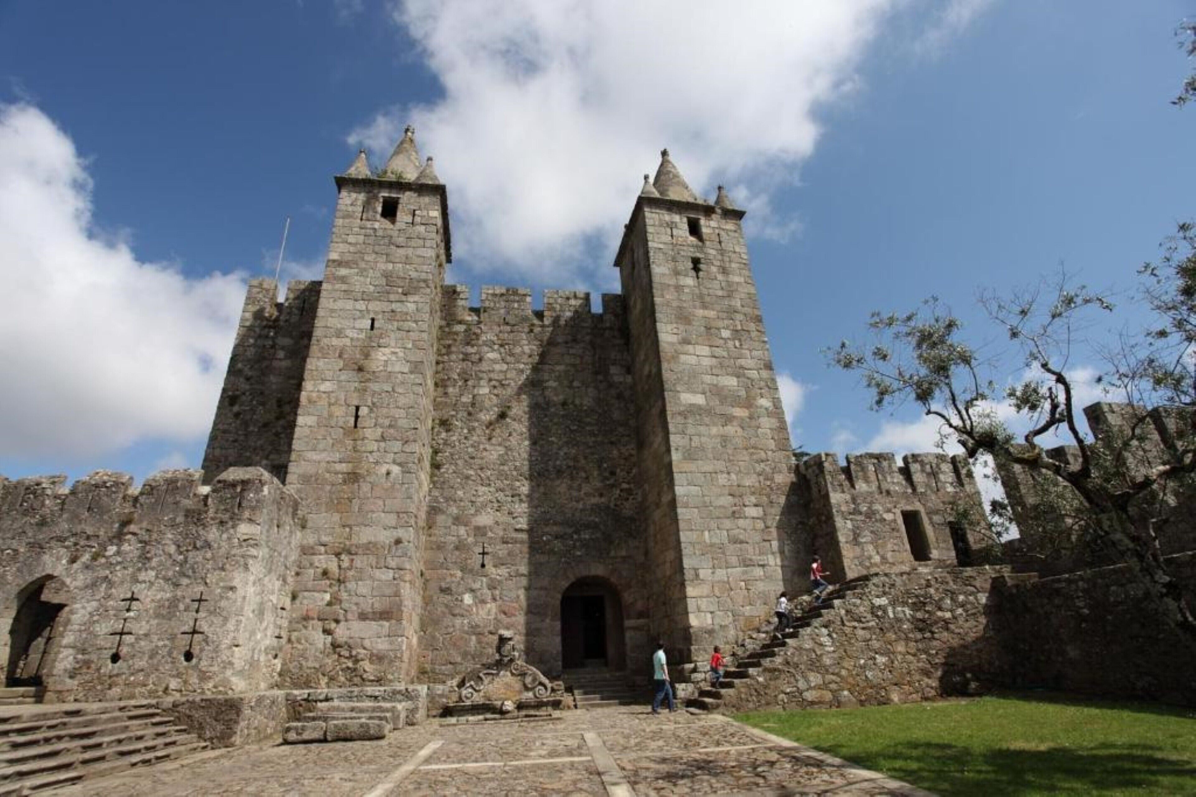 Um castelo de pedra histórico com duas torres imponentes, uma entrada central e uma muralha ao redor.