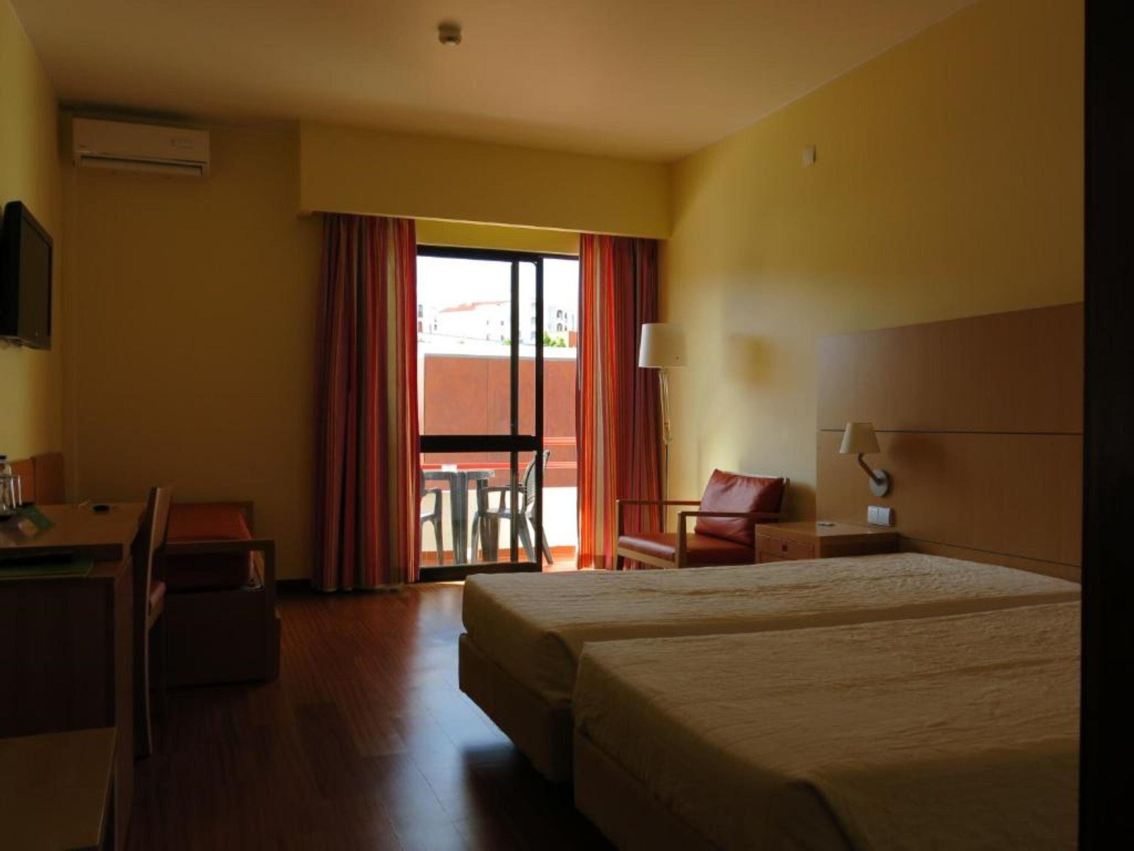 Quarto de hotel com cama, escrivaninha, cadeira, luminária e vista para o exterior.
