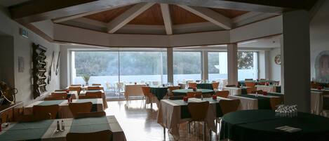 Uma sala de jantar ampla com várias mesas e cadeiras, grandes janelas com vista para o exterior e teto alto com vigas aparentes.