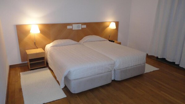 Cama de casal com duas almofadas e cabeceira de madeira. Há duas mesas de cabeceira com abajures.
