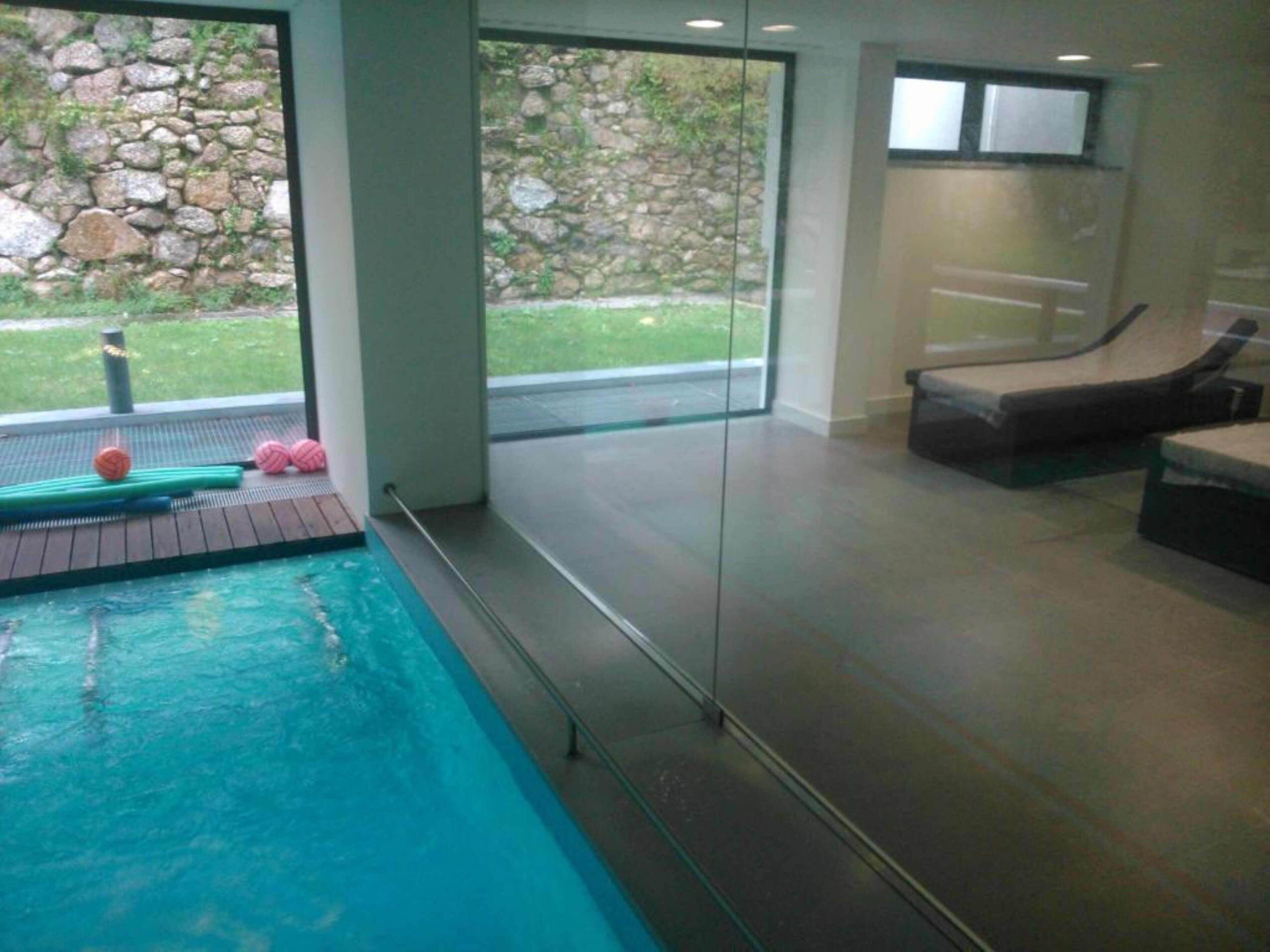 Piscina interior moderna com paredes de vidro transparente, parede de pedra no exterior e área de descanso com televisão.