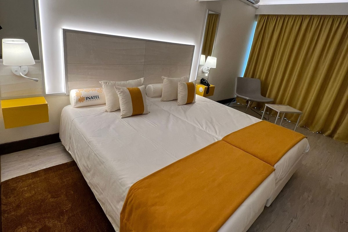 Quarto de hotel moderno com uma cama grande, uma mesinha de cabeceira, uma luminária e um espelho.