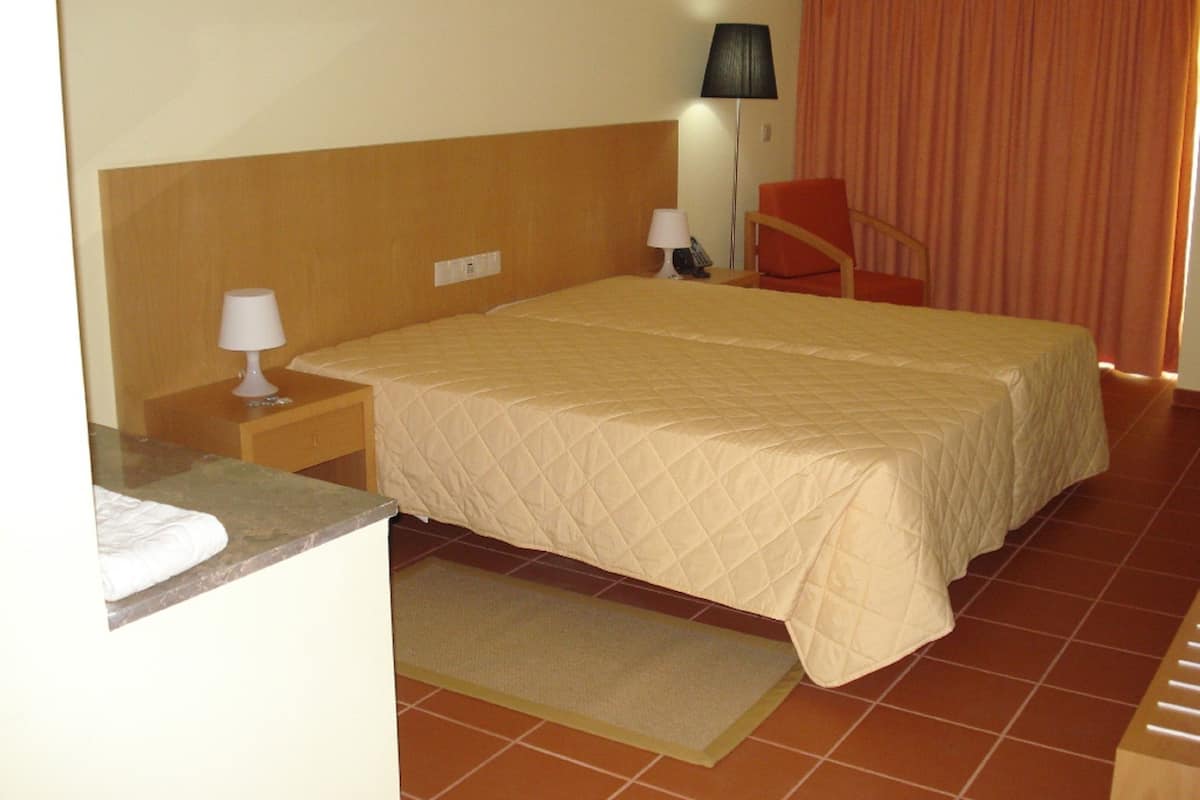 Quarto de hotel com cama, mesa de cabeceira, abajur, cadeira e janela com cortinas.