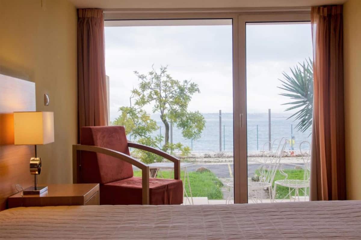 Quarto de hotel com uma cama grande, uma cadeira, uma mesinha de cabeceira e vista para a praia e o oceano através de uma porta de correr de vidro.