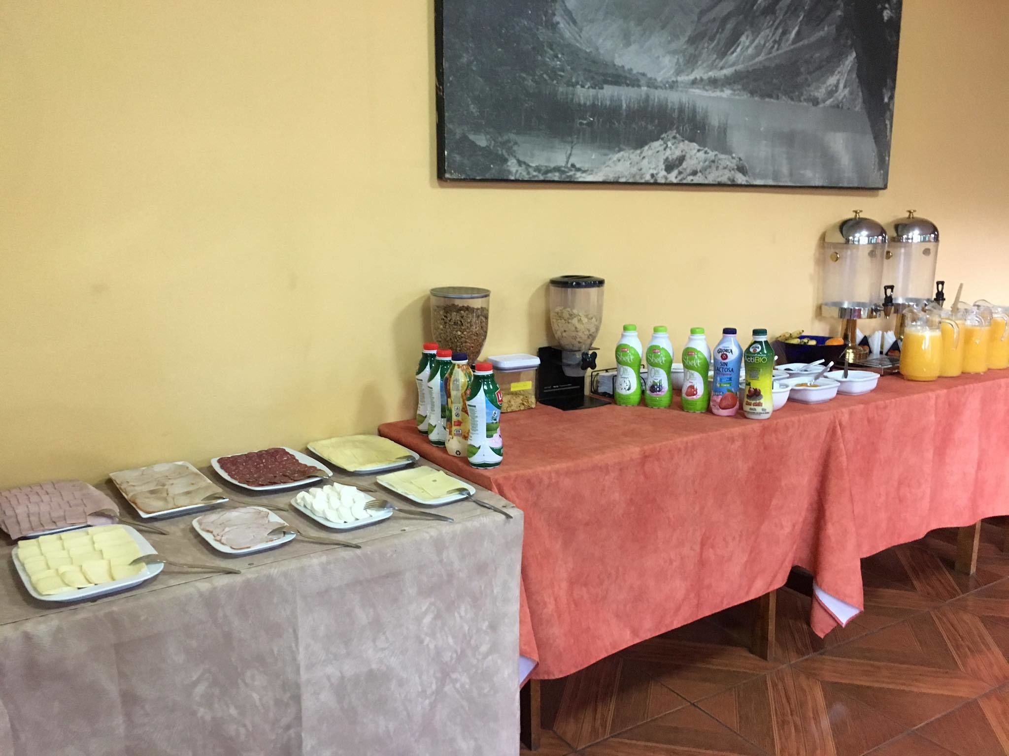 Um buffet de café da manhã com diversos alimentos e bebidas dispostos sobre uma mesa, com uma parede amarela e uma pintura em preto e branco acima.