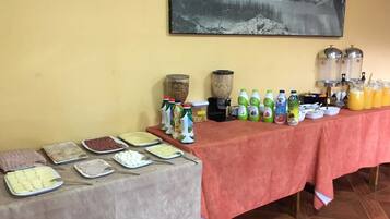Um buffet de café da manhã com diversos alimentos e bebidas dispostos sobre uma mesa, com uma parede amarela e uma pintura em preto e branco acima.