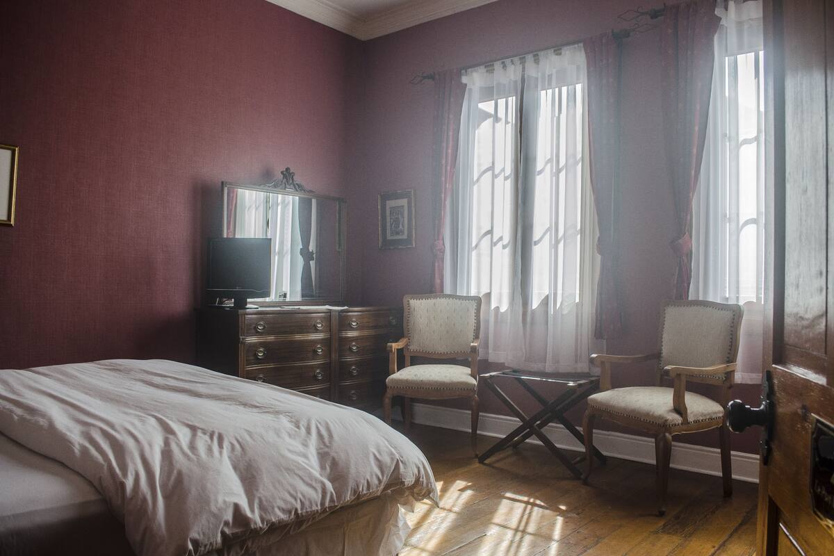 Quarto com cama, cômoda, duas cadeiras, espelho e janela com cortinas.