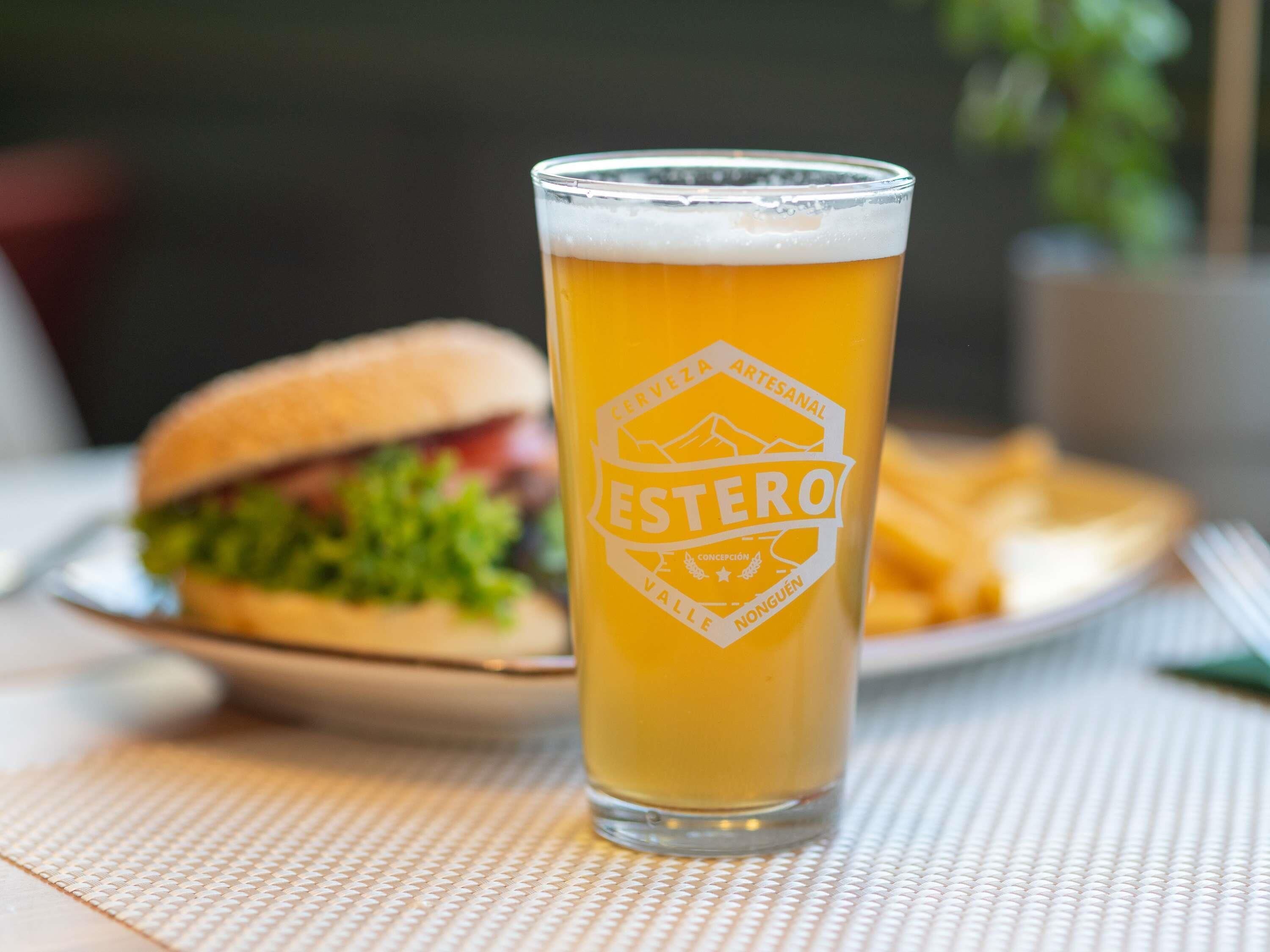Um copo de cerveja com etiqueta, um hambúrguer e batatas fritas em cima de uma mesa.