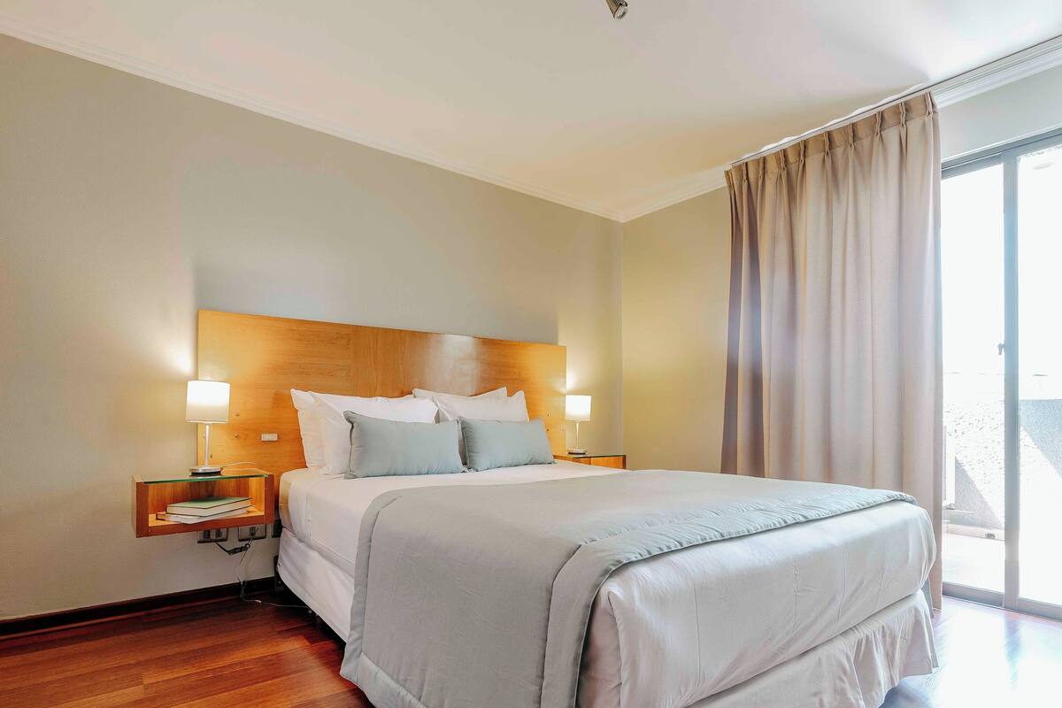 Quarto de hotel com uma cama grande, duas mesas de cabeceira com abajures, um painel de madeira na cabeceira e uma janela grande com cortinas.