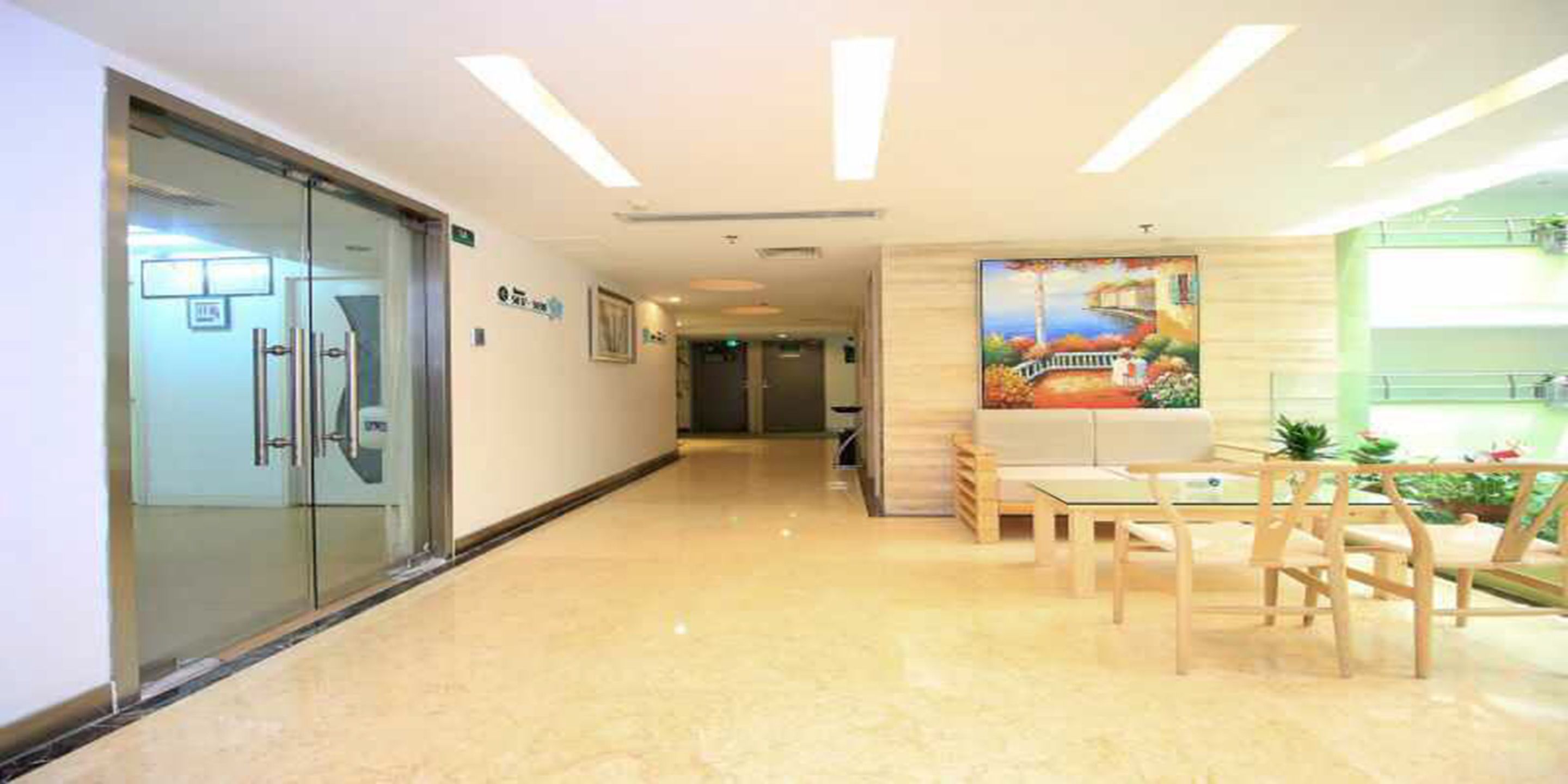 Um corredor de hotel com entrada por porta de vidro, uma área de refeições com mesa e cadeiras, e um quadro na parede.