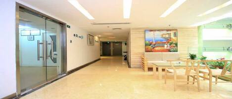 Um corredor de hotel com entrada por porta de vidro, uma área de refeições com mesa e cadeiras, e um quadro na parede.