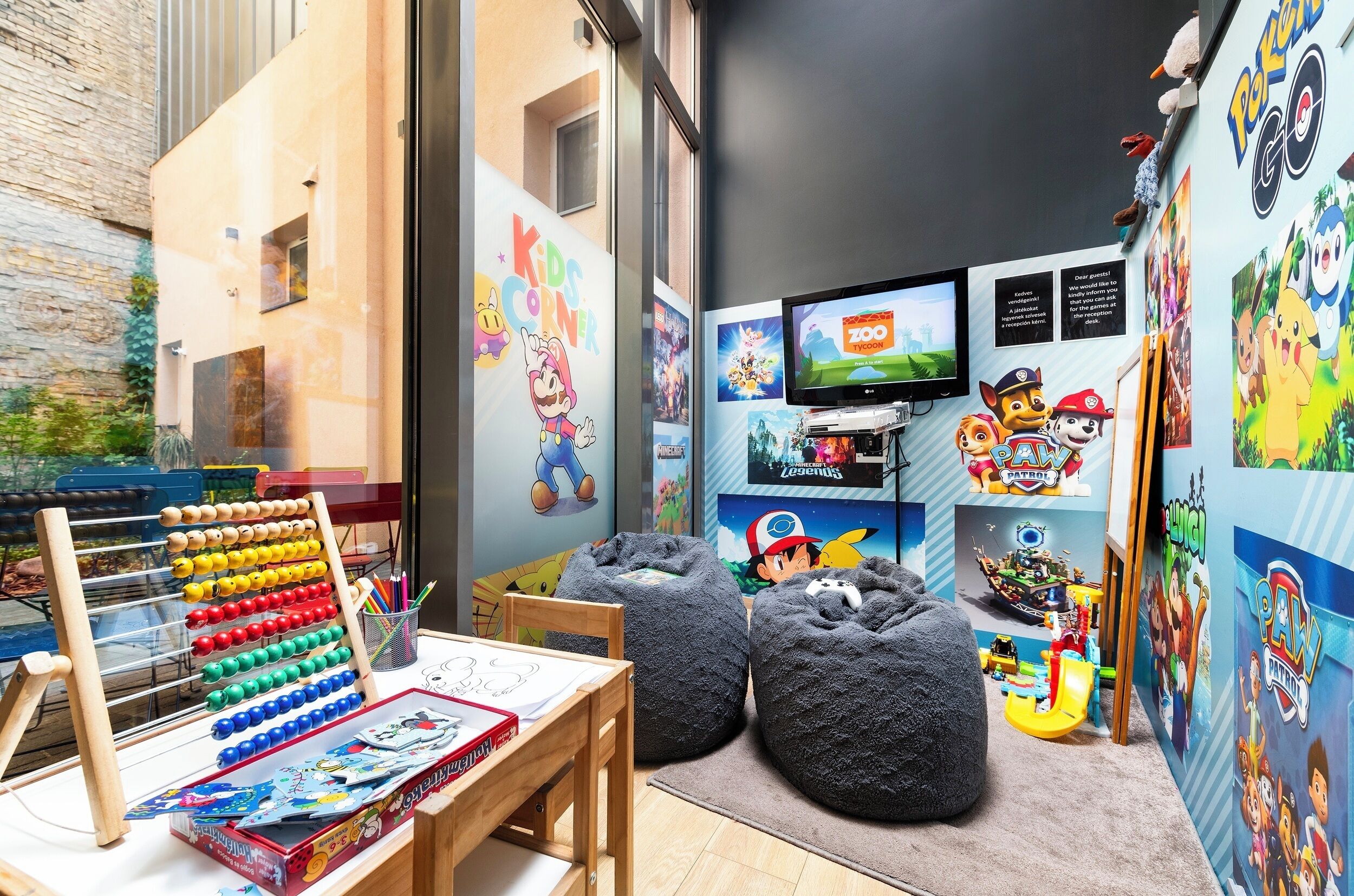 Espaço infantil com um ábaco colorido, poltronas de grãos e diversos consoles de jogos.
