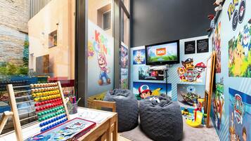 Espaço infantil com um ábaco colorido, poltronas de grãos e diversos consoles de jogos.