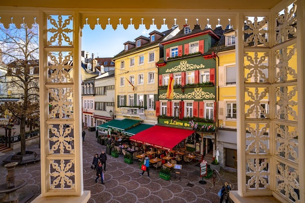 Uma praça de paralelepípedos com mesas ao ar livre, um café e um edifício com toldo verde e persianas vermelhas.