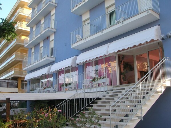 Um hotel com vários andares, varandas, área de estar externa e escadaria que leva à entrada.