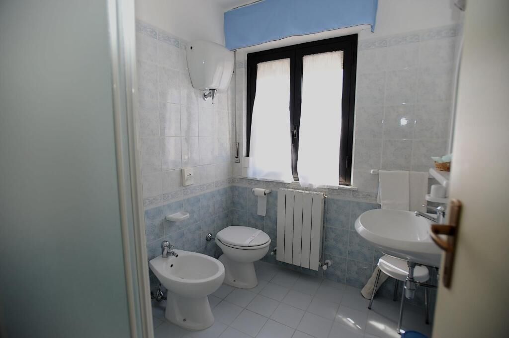 Casa de banho com sanita, bidé e lavatório, revestida a mosaicos em tons claros e escuros, com janela e radiador.