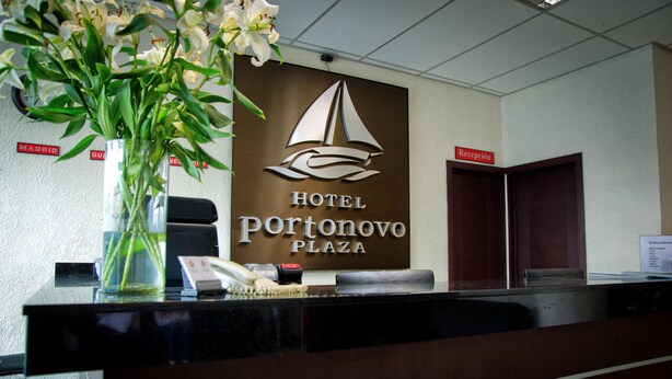 Bancada de recepção no Hotel Portonovo Plaza, com um logotipo, um vaso de flores e uma placa indicando a área de recepção.
