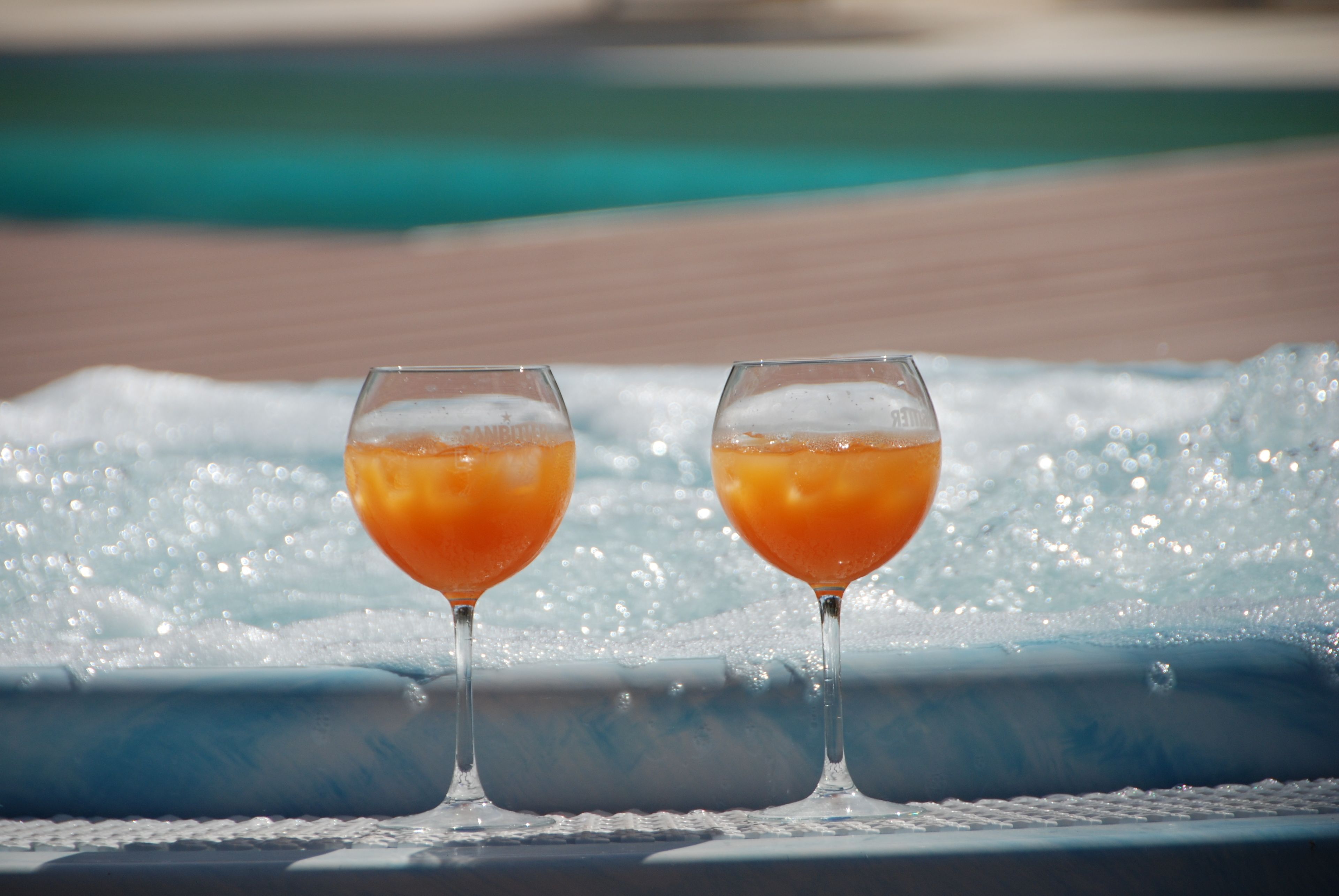 Dois copos de bebida de laranja sobre uma mesa à beira da piscina, com a piscina ao fundo.