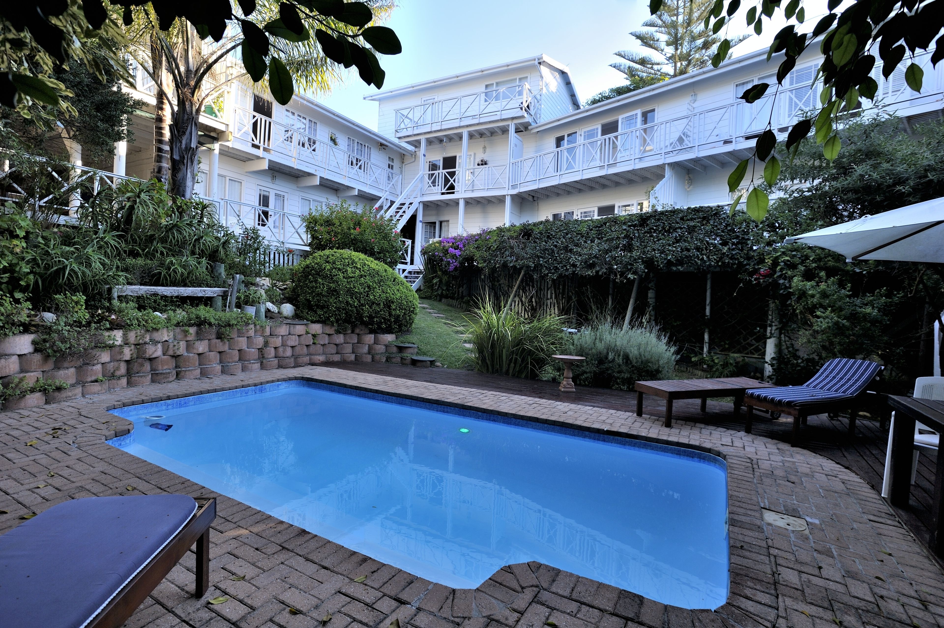 Piscina com deck de tijolos, rodeada por um jardim com diversas plantas e um banco.