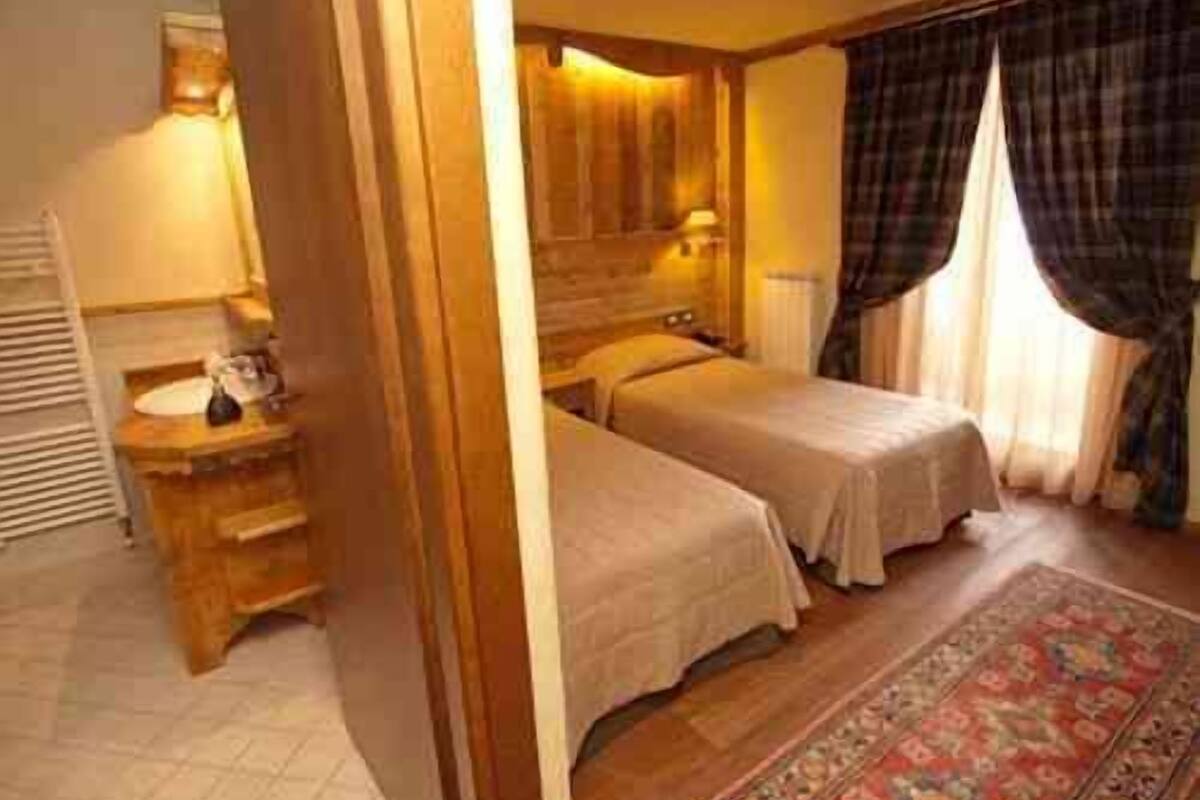 Quarto com duas camas, cômoda de madeira, mesa de cabeceira e uma janela grande com cortinas.