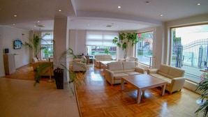 Um hall amplo com piso de madeira, sofás brancos, mesa de centro e grandes vasos de plantas.