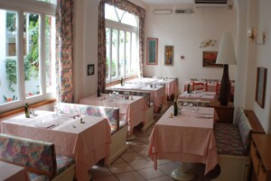 Uma sala de jantar com várias mesas e bancos, janelas amplas com cortinas e piso em azulejos.