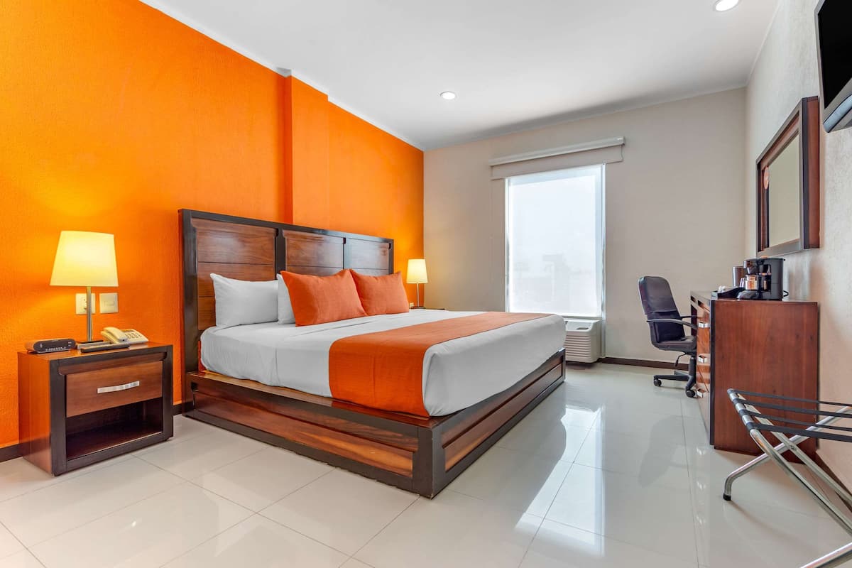 Quarto de hotel com uma cama grande, duas mesas de cabeceira com abajures, uma escrivaninha com cadeira e uma grande janela com vista para a cidade.