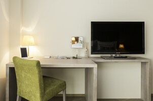 Quarto de hotel moderno com escrivaninha, cadeira, luminária, espelho e televisão de tela plana.