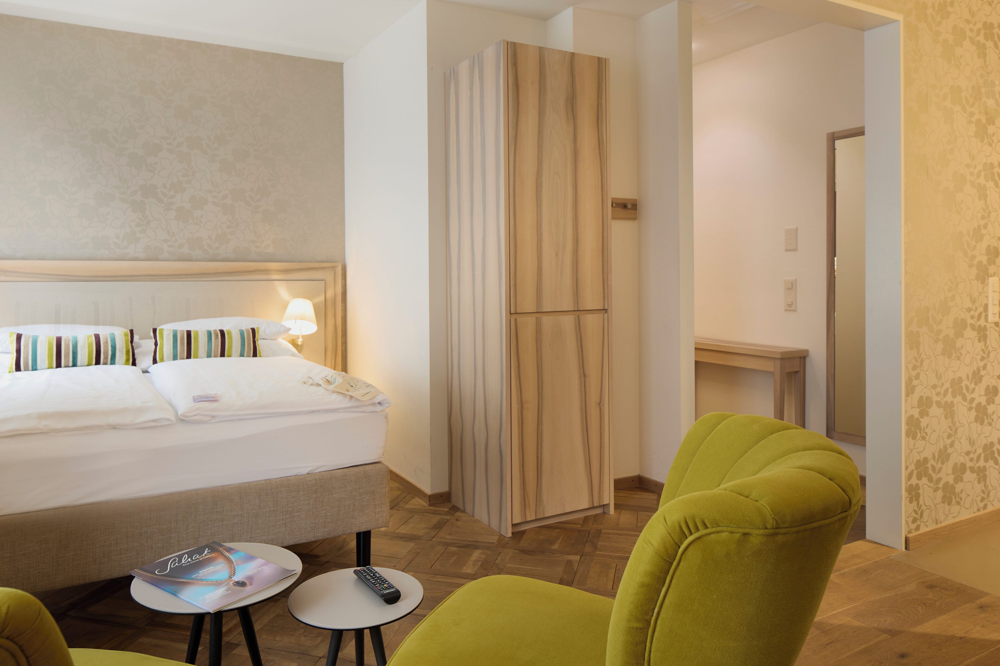 Quarto de hotel com uma cama, uma mesinha com um controle remoto e uma poltrona verde.