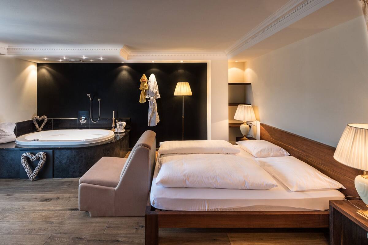 Quarto de hotel moderno com uma cama grande, área de estar, banheira e toucador com espelho.