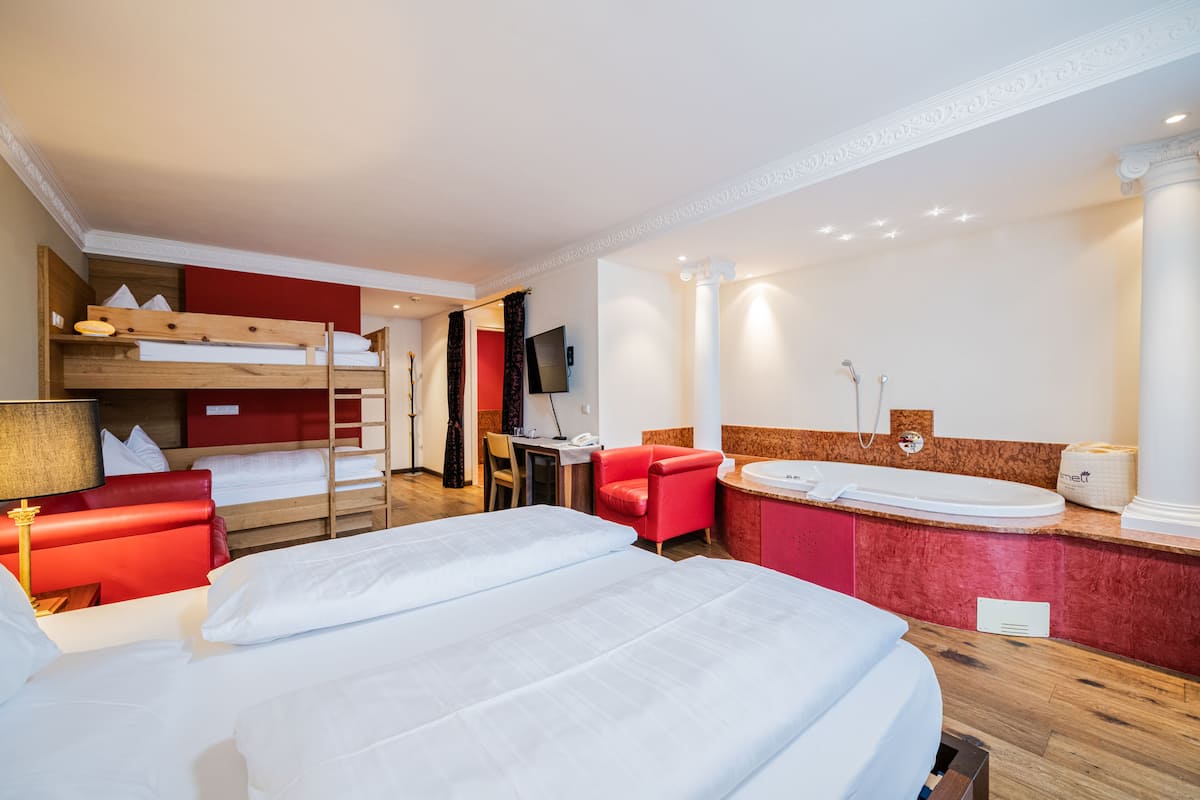 Quarto de hotel com cama beliche, uma poltrona vermelha, escrivaninha com computador, banheira grande e televisão.