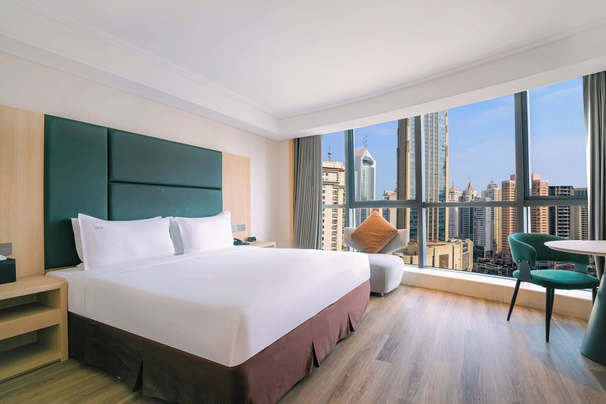Quarto de hotel com uma cama grande, uma mesa pequena, uma cadeira e vista para a cidade.
