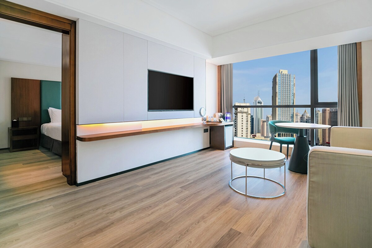 Quarto de hotel moderno com televisão de tela plana, escrivaninha de madeira, mesa redonda e vista para a paisagem urbana.