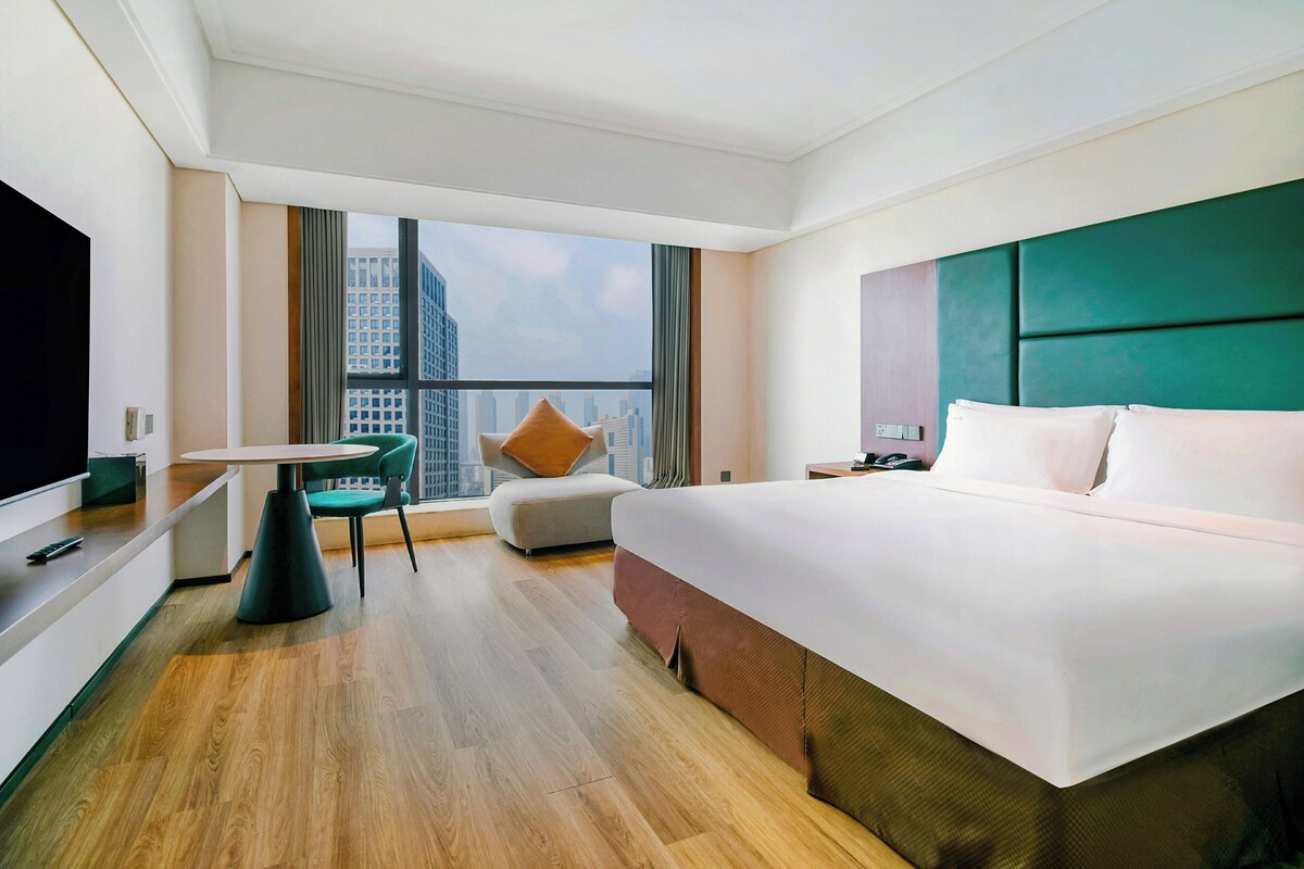 Quarto de hotel com uma cama grande, televisão, escrivaninha com cadeira, vista da cidade por grandes janelas e paisagem urbana visível.