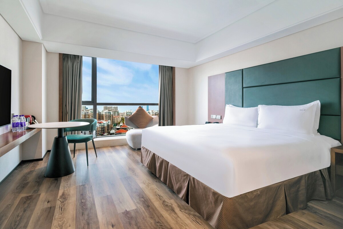 Quarto de hotel com uma cama grande, uma escrivaninha com cadeira, vista da cidade e paisagem urbana visível pela janela.