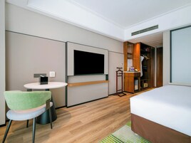 Quarto de hotel com cama, televisão presa na parede, mesa redonda pequena, cadeira verde, guarda-roupa e chão de madeira.