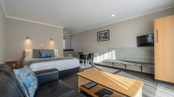Quarto de hotel moderno com cama, sofá, mesa de centro de madeira, escrivaninha, cadeira, televisão e guarda-roupa.