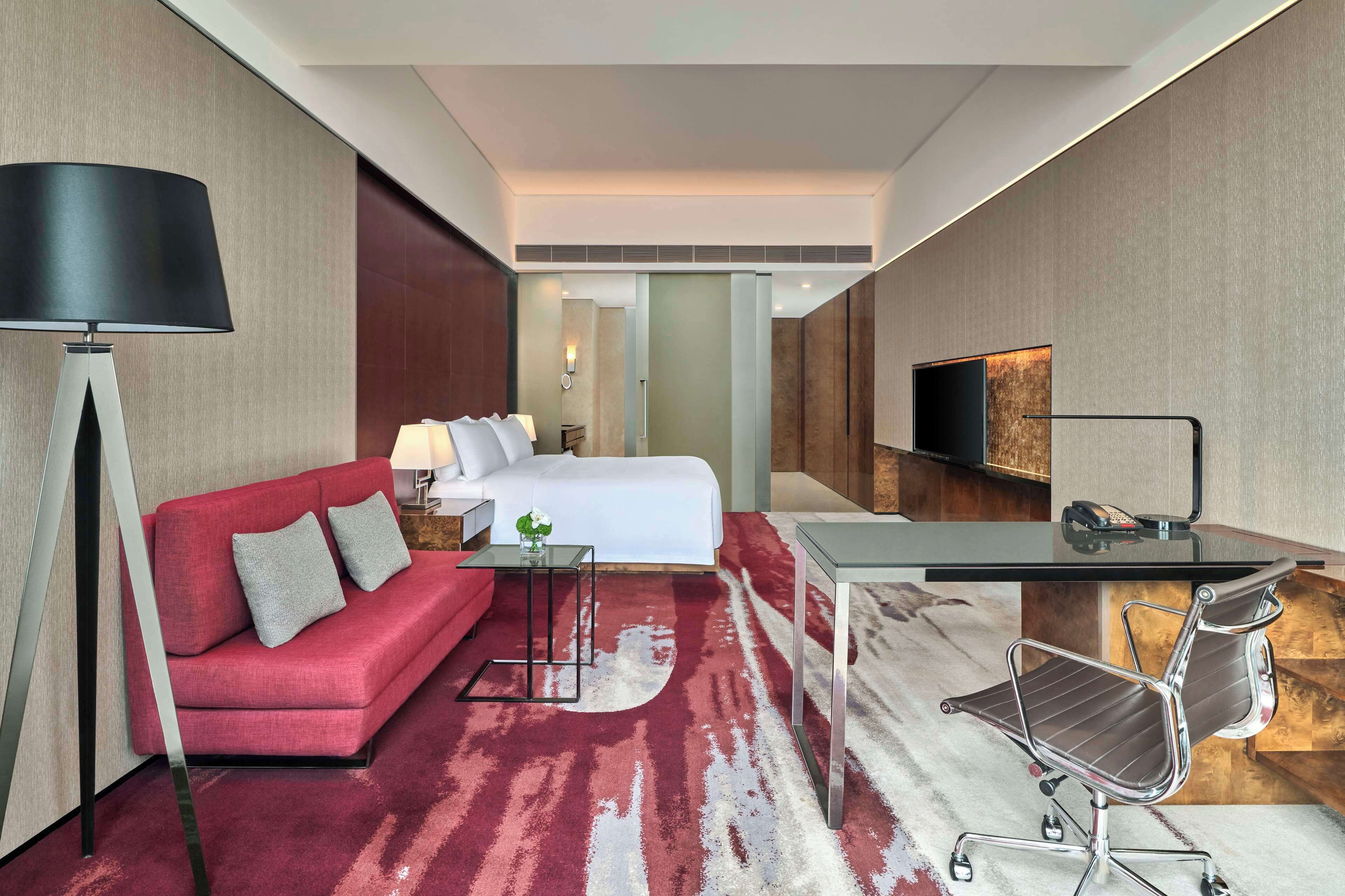 Quarto de hotel com sofá vermelho, escrivaninha com cadeira, cama, televisão e abajur.