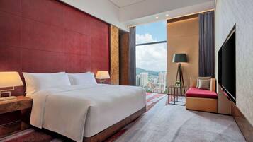 Quarto de hotel moderno com cama grande, televisão, área de estar com vista e paisagem urbana visível pela janela.