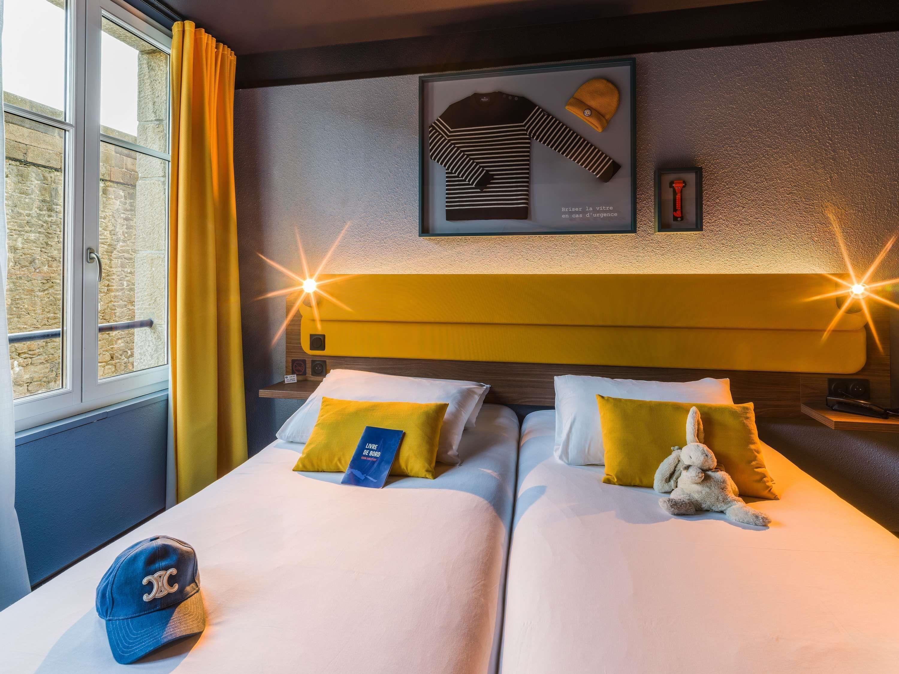 Quarto de hotel com duas camas, janela com cortinas, cabeceira com iluminação embutida, um objeto emoldurado na parede e uma pequena mesa com um abajur e um brinquedo de pelúcia.