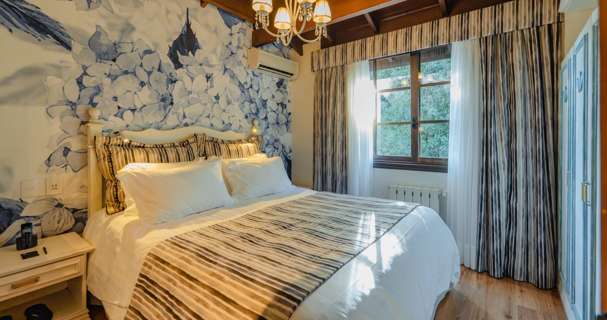 Quarto com cama grande, roupa de cama listrada, parede florida, janela com cortinas e lustre.