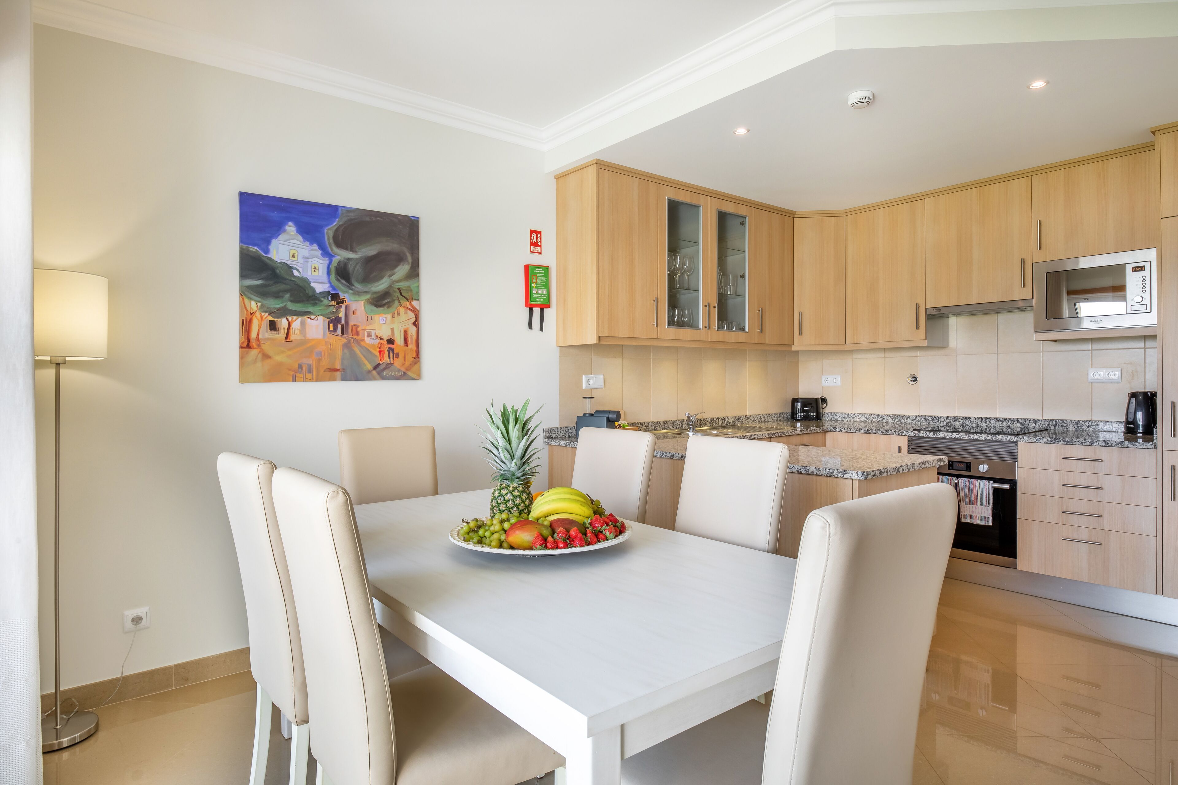 Cozinha moderna com armários de madeira, micro-ondas e área de jantar com mesa e cadeiras brancas.