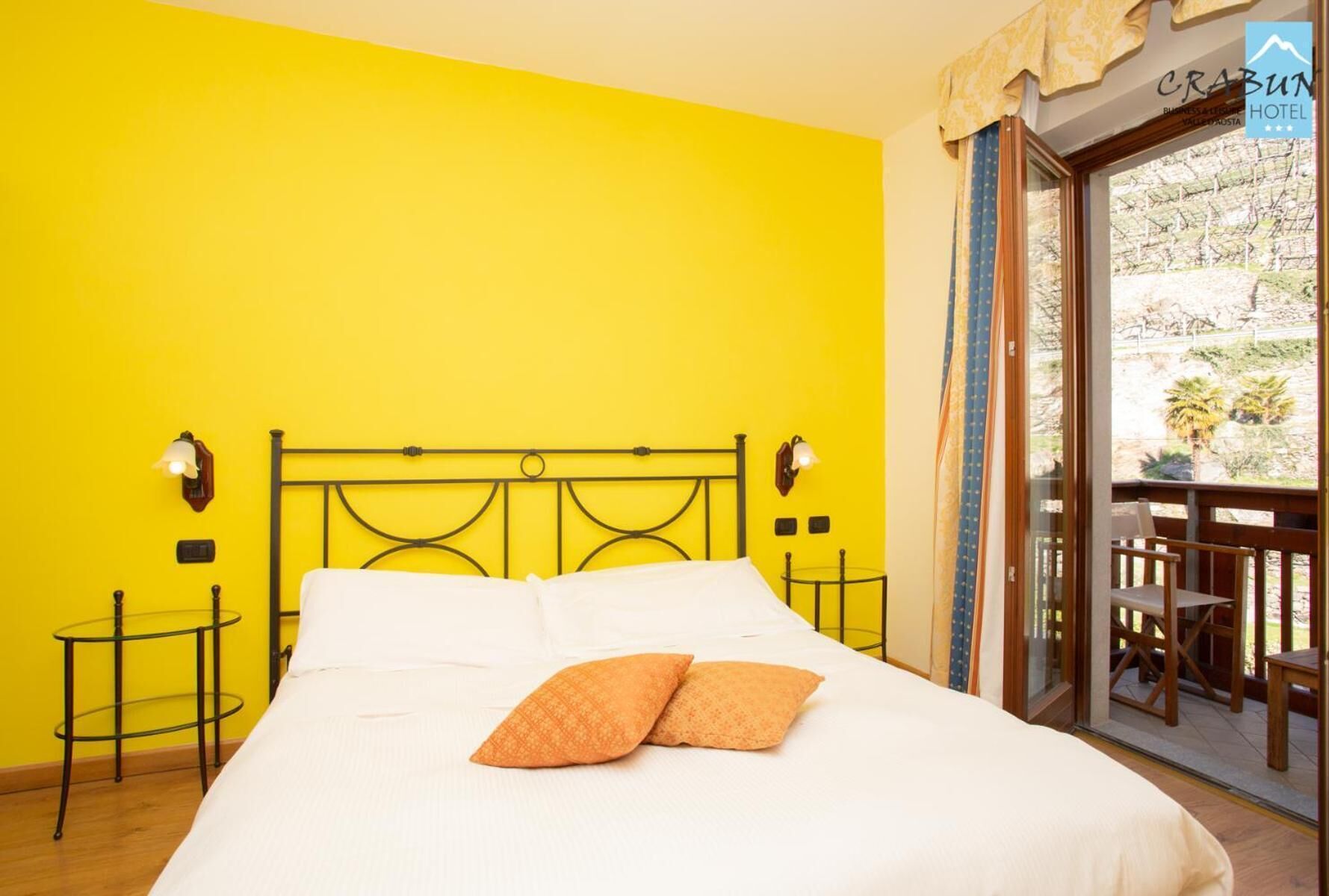 Quarto amarelo e iluminado com uma cama, duas luminárias fixadas na parede e vista para uma varanda com mesa e cadeiras.