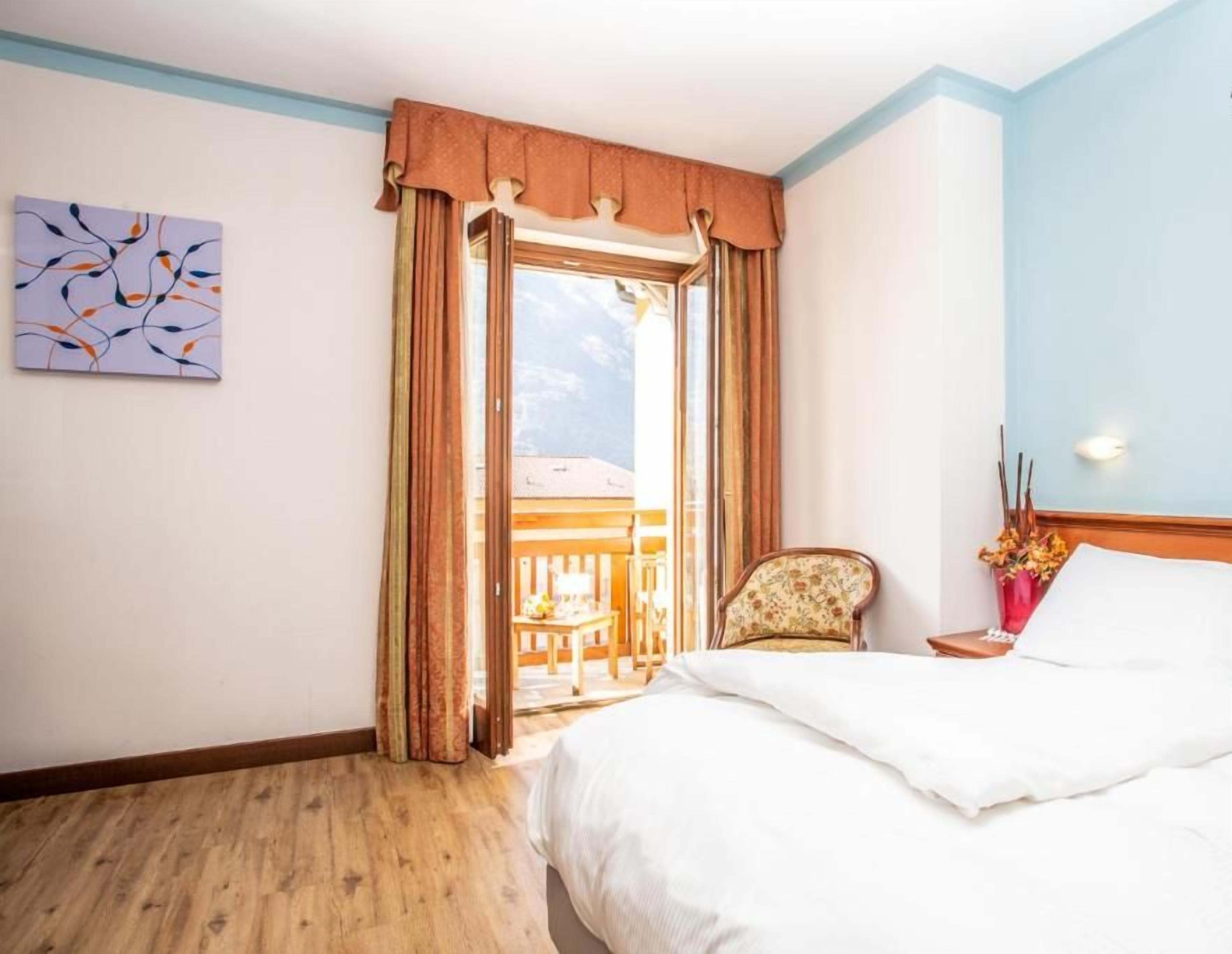 Quarto de hotel com cama, cadeira, mesa de cabeceira, janela com cortinas e um quadro na parede.