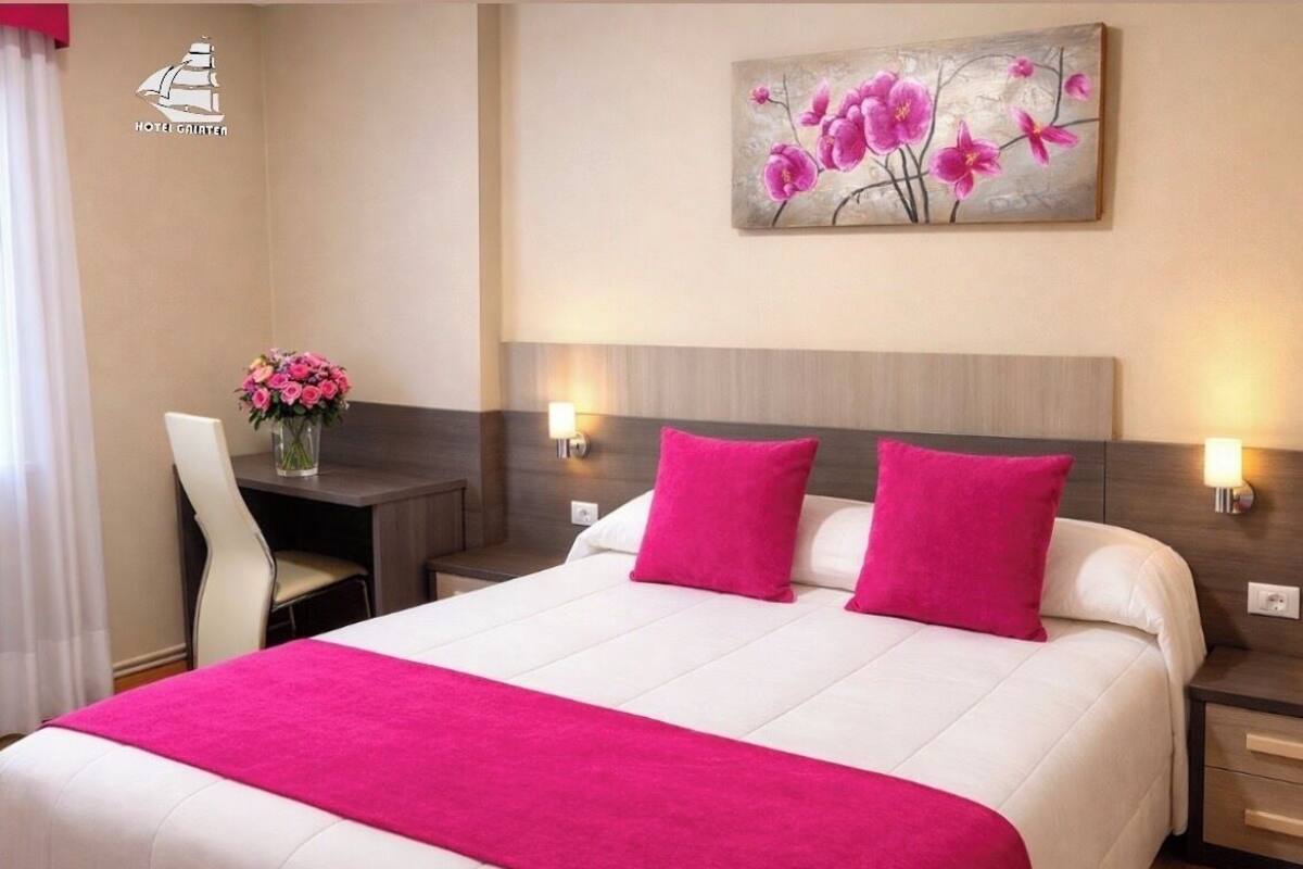 Quarto de hotel com uma cama, escrivaninha, cadeira, mesa de cabeceira e um quadro na parede.