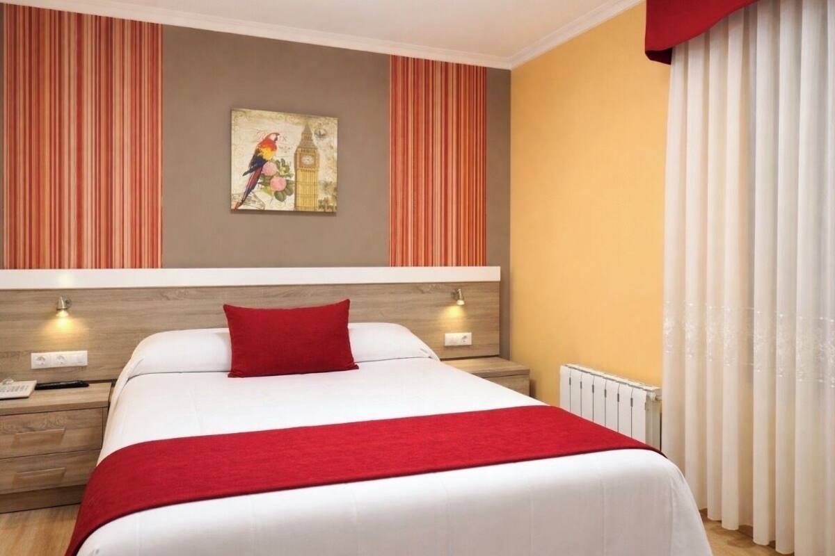 Quarto de hotel com cama, mesa de cabeceira, luminária na parede, radiador e janela com cortinas.