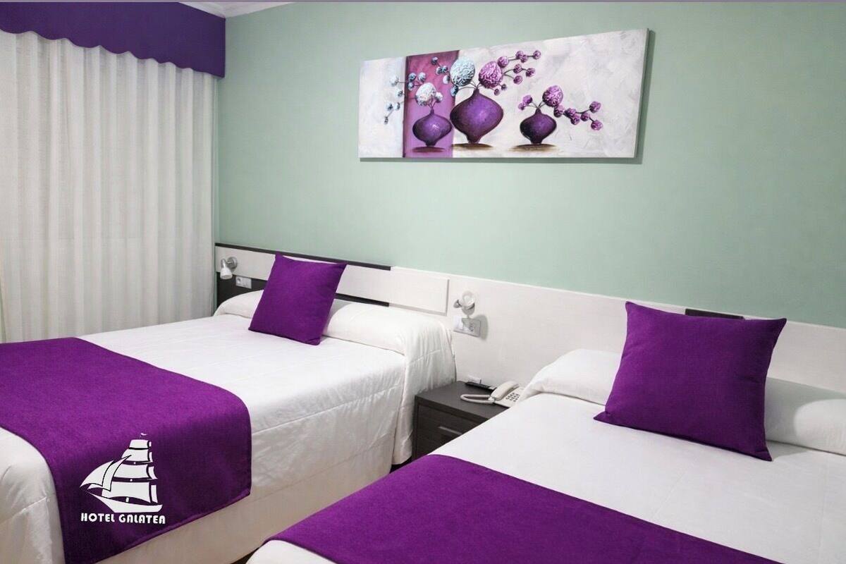 Quarto de hotel com duas camas, roupa de cama branca, detalhes em roxo, uma pintura de vasos e o logotipo no edredom.
