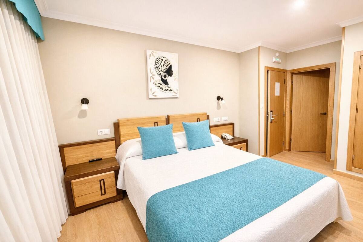 Quarto de hotel com cama, mesas de cabeceira, luminária na parede, porta e janela com cortinas.