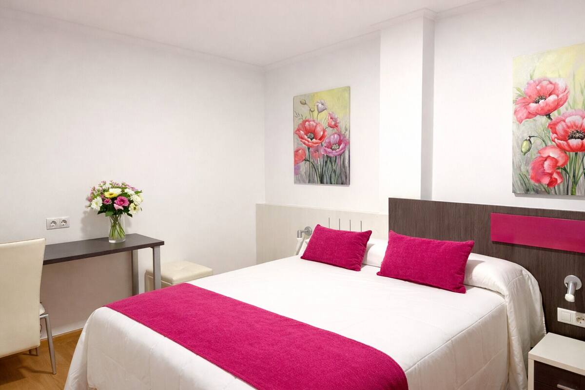 Quarto de hotel com cama, escrivaninha, cadeira e duas pinturas na parede.